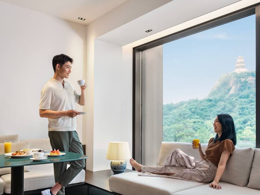 Quarto de hotel moderno com uma grande janela que oferece vista para uma montanha e um templo. Há uma área de refeições com itens de café da manhã e um abajur.