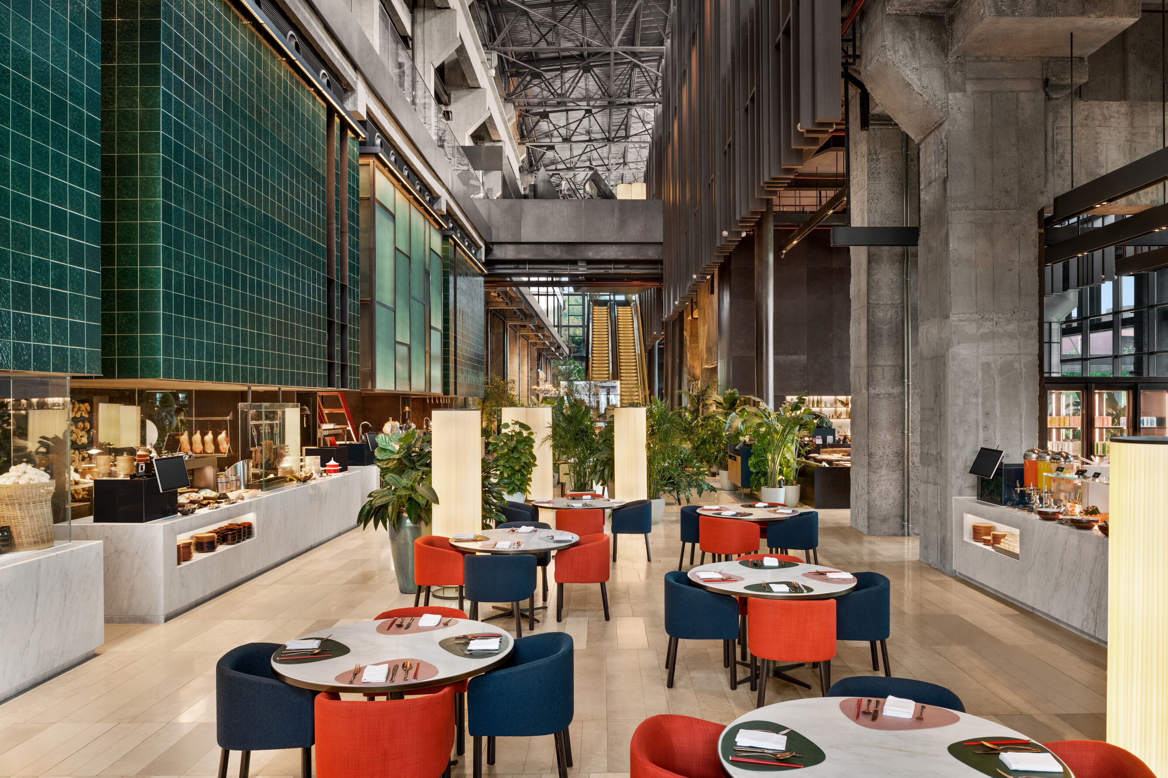 Um espaço amplo e interno com várias mesas e cadeiras, um balcão comprido com exposição de produtos e grandes janelas que permitem entrada de luz natural.