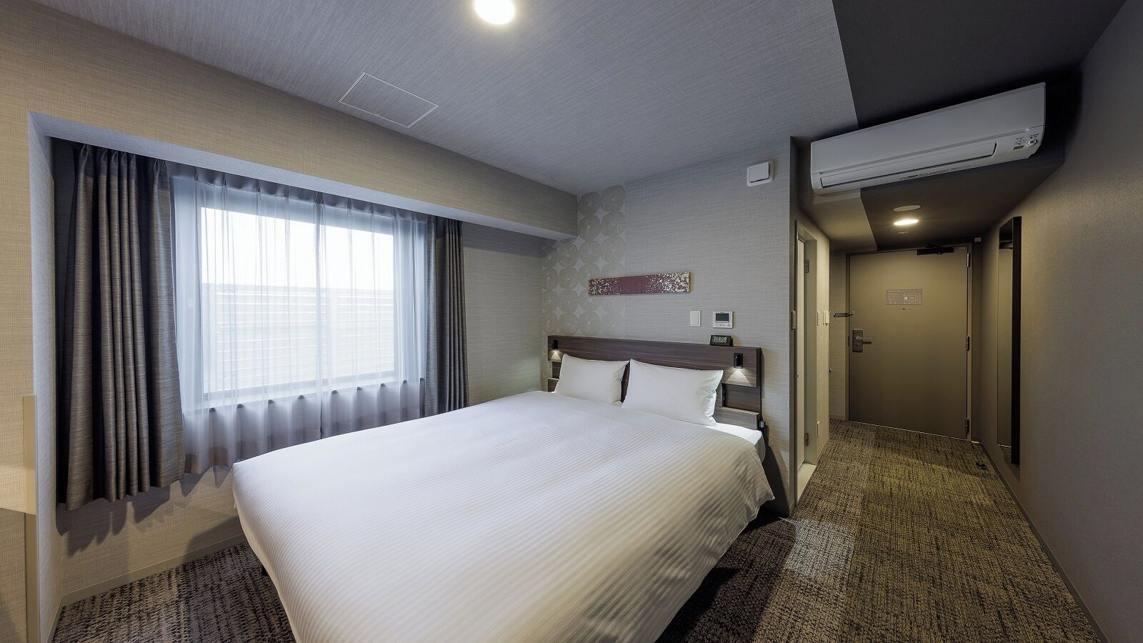 Quarto de hotel com uma cama grande, janela com cortinas, ar condicionado, relógio de parede e piso coberto de carpete.