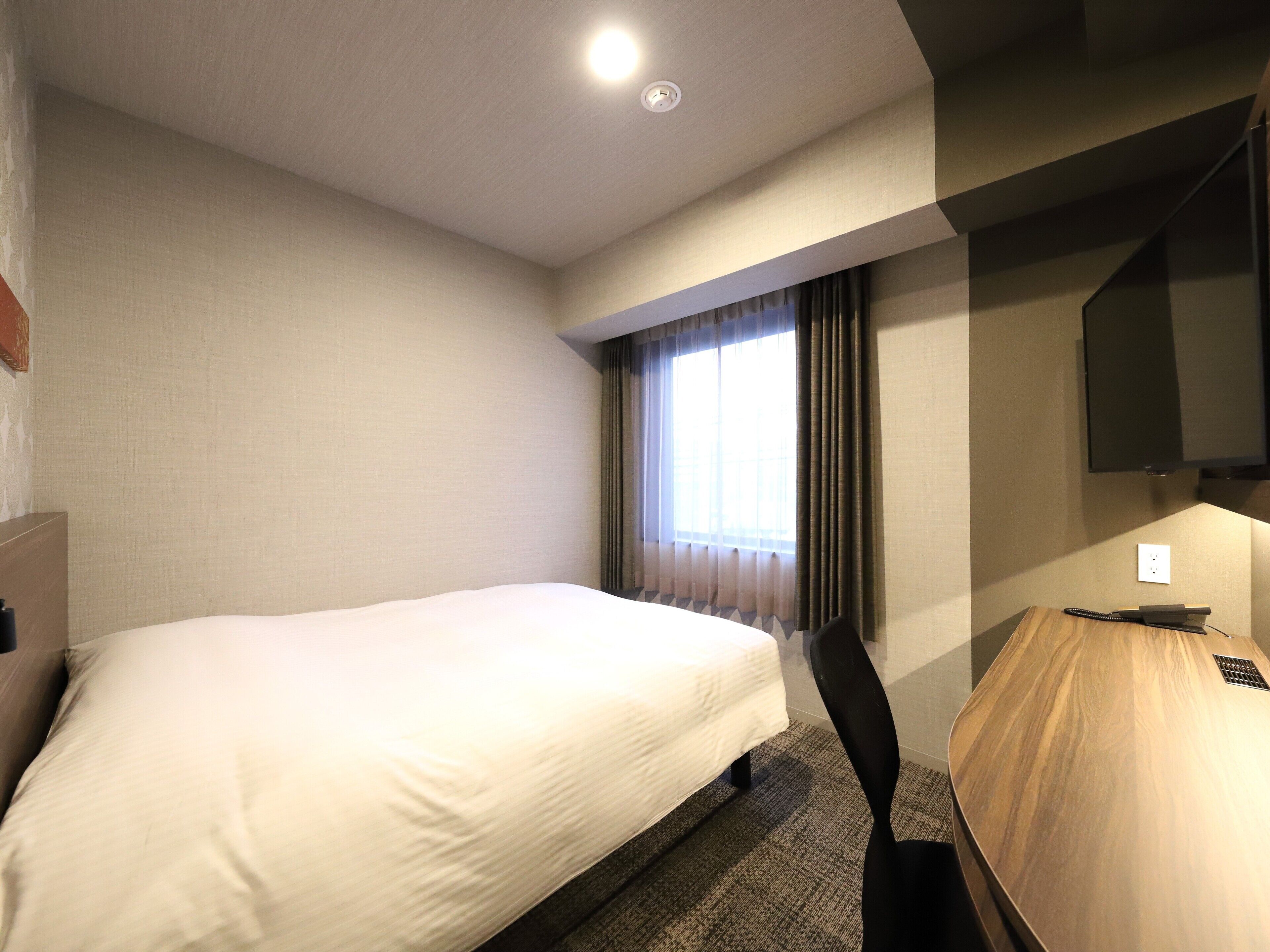 Quarto de hotel com cama, escrivaninha, cadeira, televisão e janela com cortinas.