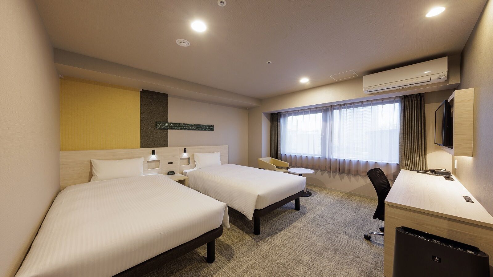 Quarto de hotel com duas camas, uma escrivaninha, uma cadeira, uma mesinha e ar condicionado, além de uma televisão.
