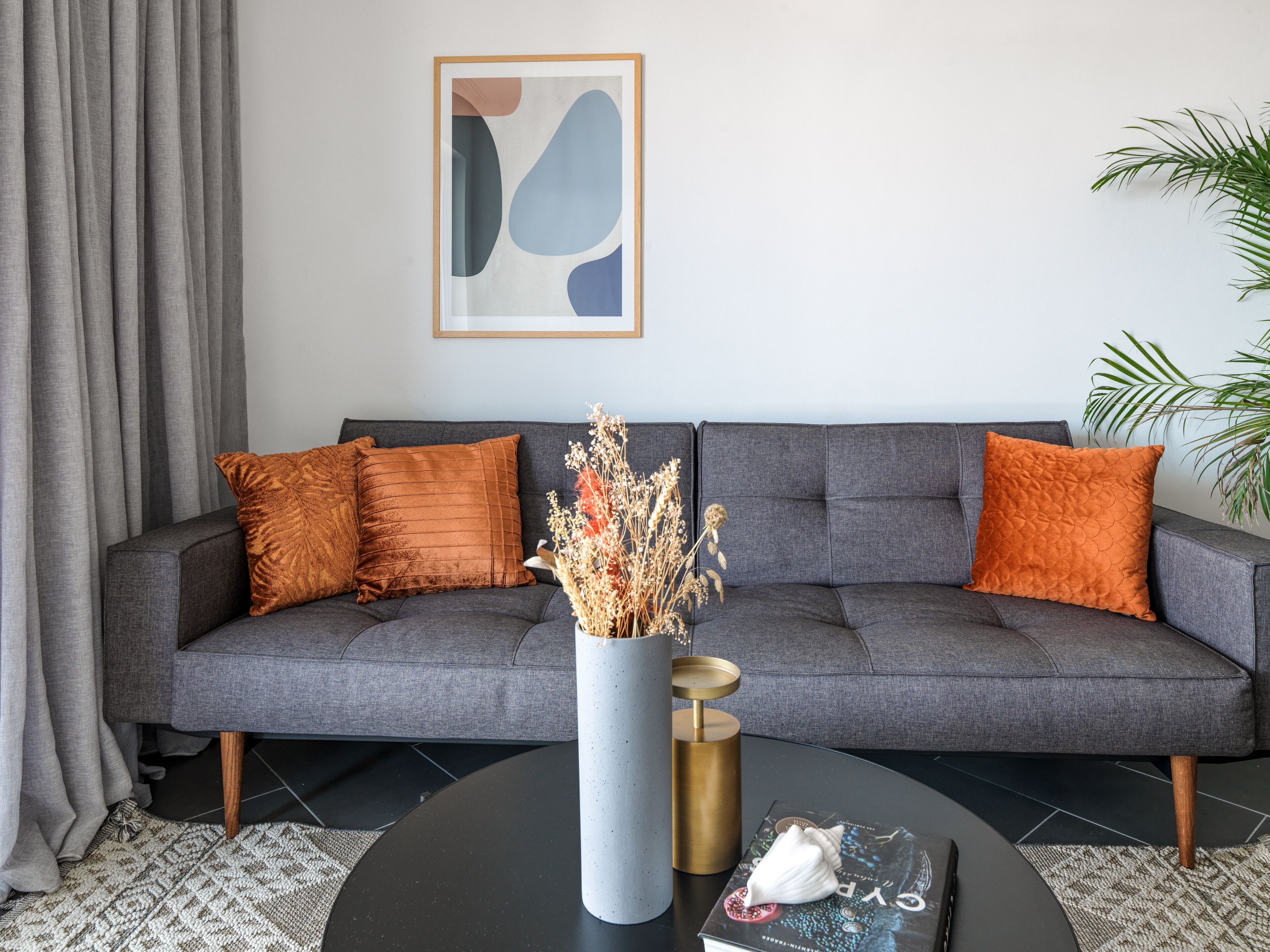 Uma sala de estar moderna com um sofá cinzento, almofadas laranja, um vaso com flores secas e uma pintura abstrata emoldurada na parede.