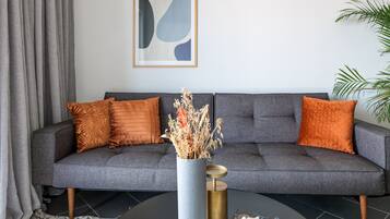 Uma sala de estar moderna com um sofá cinzento, almofadas laranja, um vaso com flores secas e uma pintura abstrata emoldurada na parede.