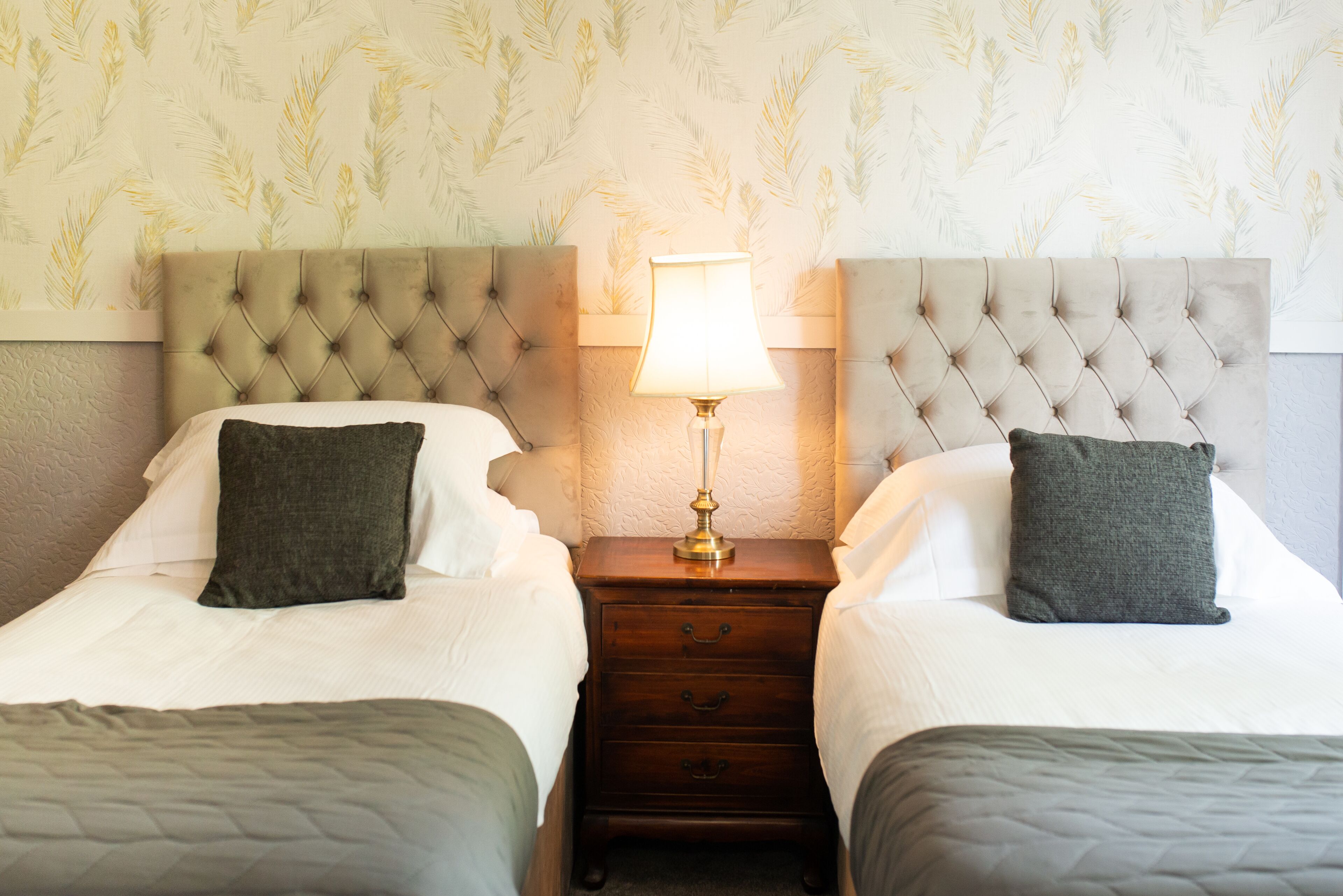 Quarto de hotel com duas camas, uma mesa de cabeceira com abajur e papel de parede estampado.