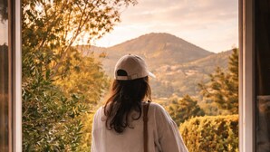 Uma pessoa apoiada no parapeito de uma janela, com uma mala, observando uma paisagem montanhosa.