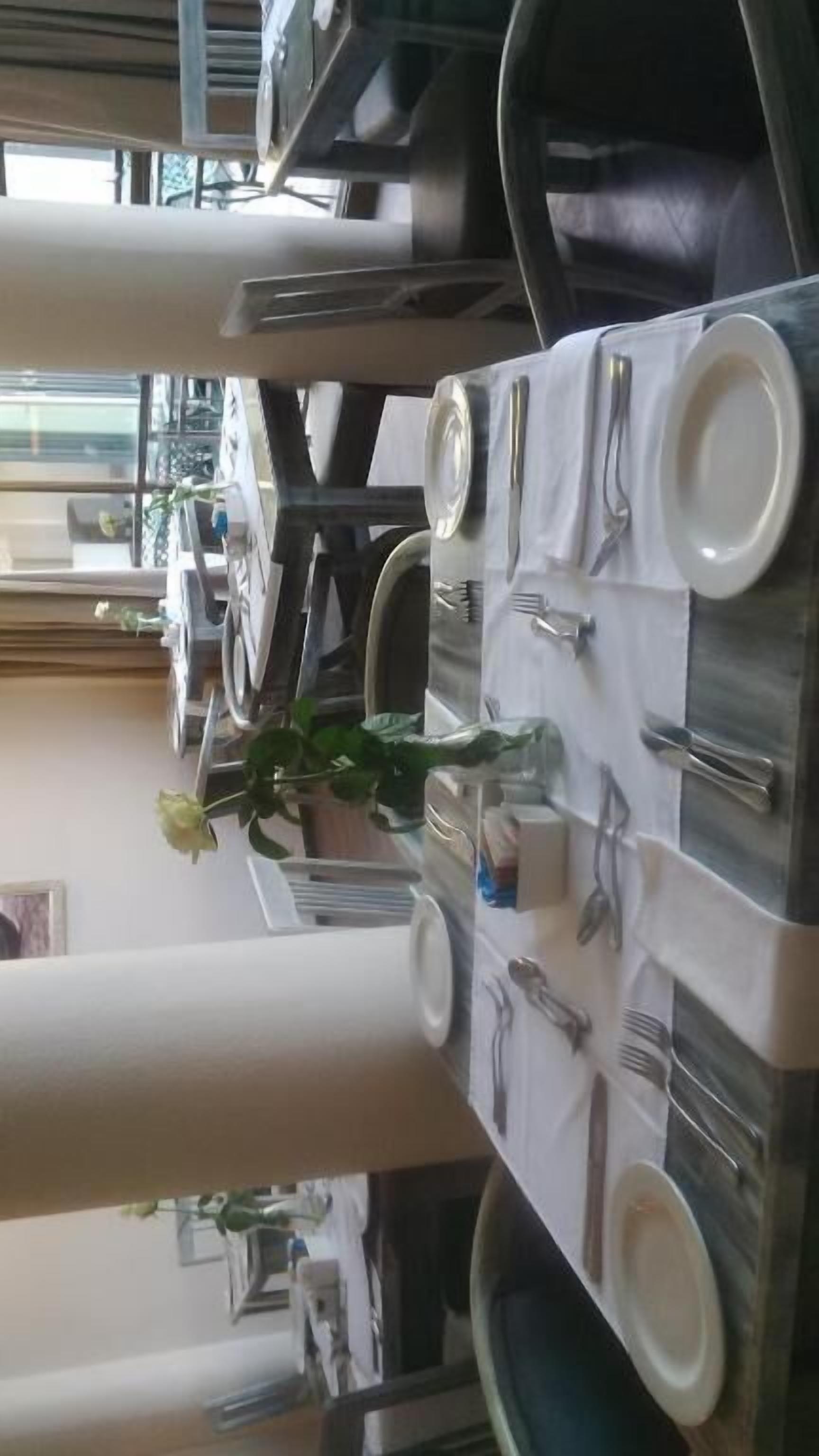 Uma sala de jantar com uma mesa comprida posta para várias pessoas, incluindo pratos, talheres e um vaso com uma única flor.