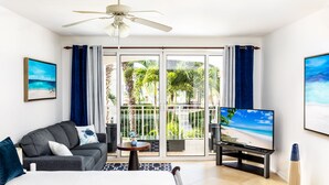 Quarto de hotel moderno com cama, sofá, televisão em um suporte, ventilador de teto e varanda com vista para palmeiras e uma praia.