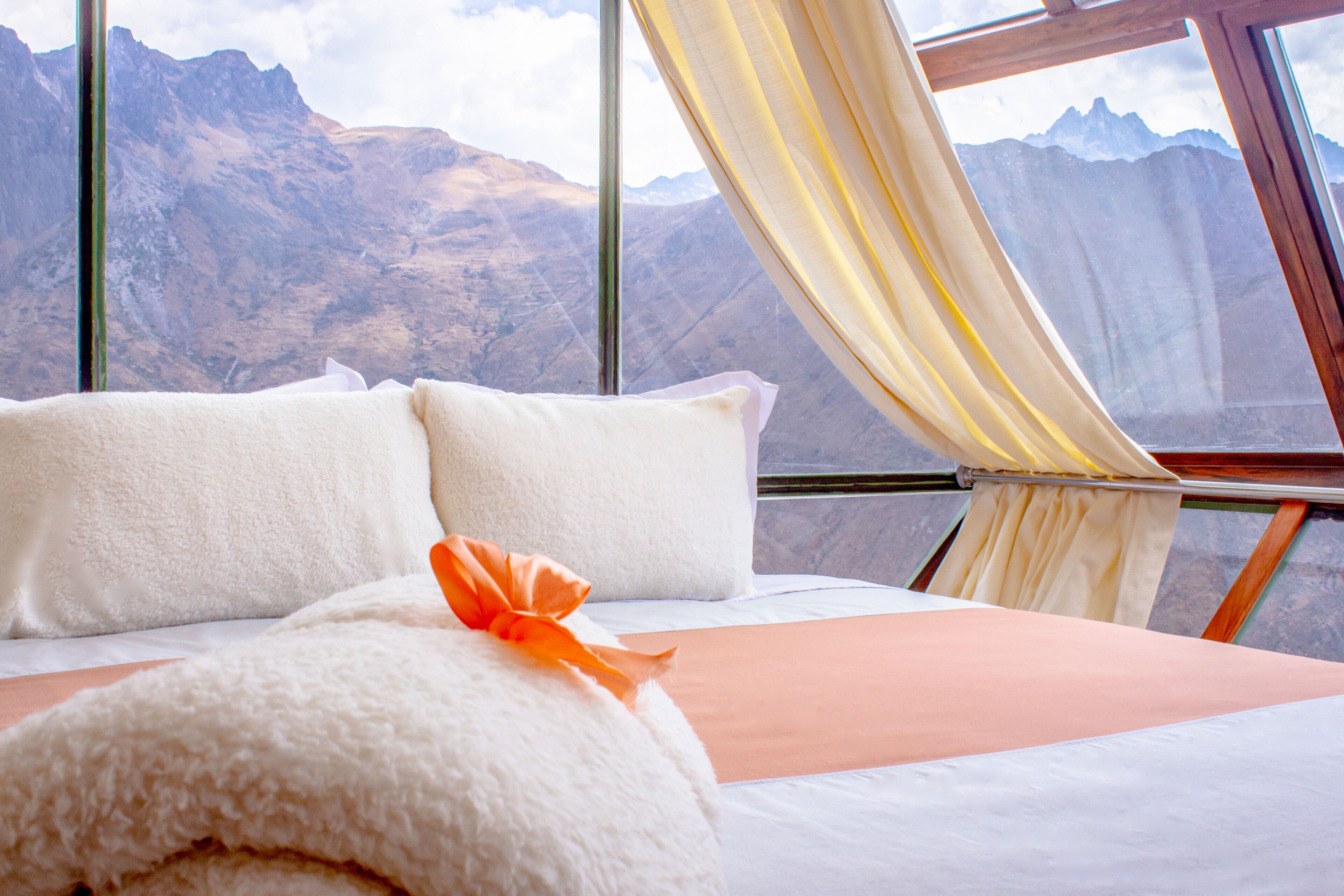 Uma cama com almofadas e manta brancas, uma janela com cortinas e vista para a montanha.