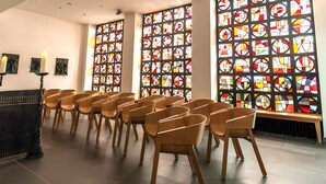 Interior de igreja com cadeiras de madeira dispostas em fileiras, um banco de madeira e uma janela de vitrais com padrões geométricos.