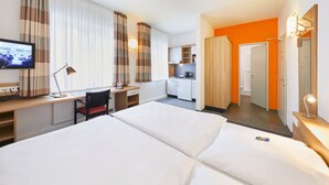 Quarto de hotel com cama, escrivaninha, cadeira, televisão e cozinha compacta.