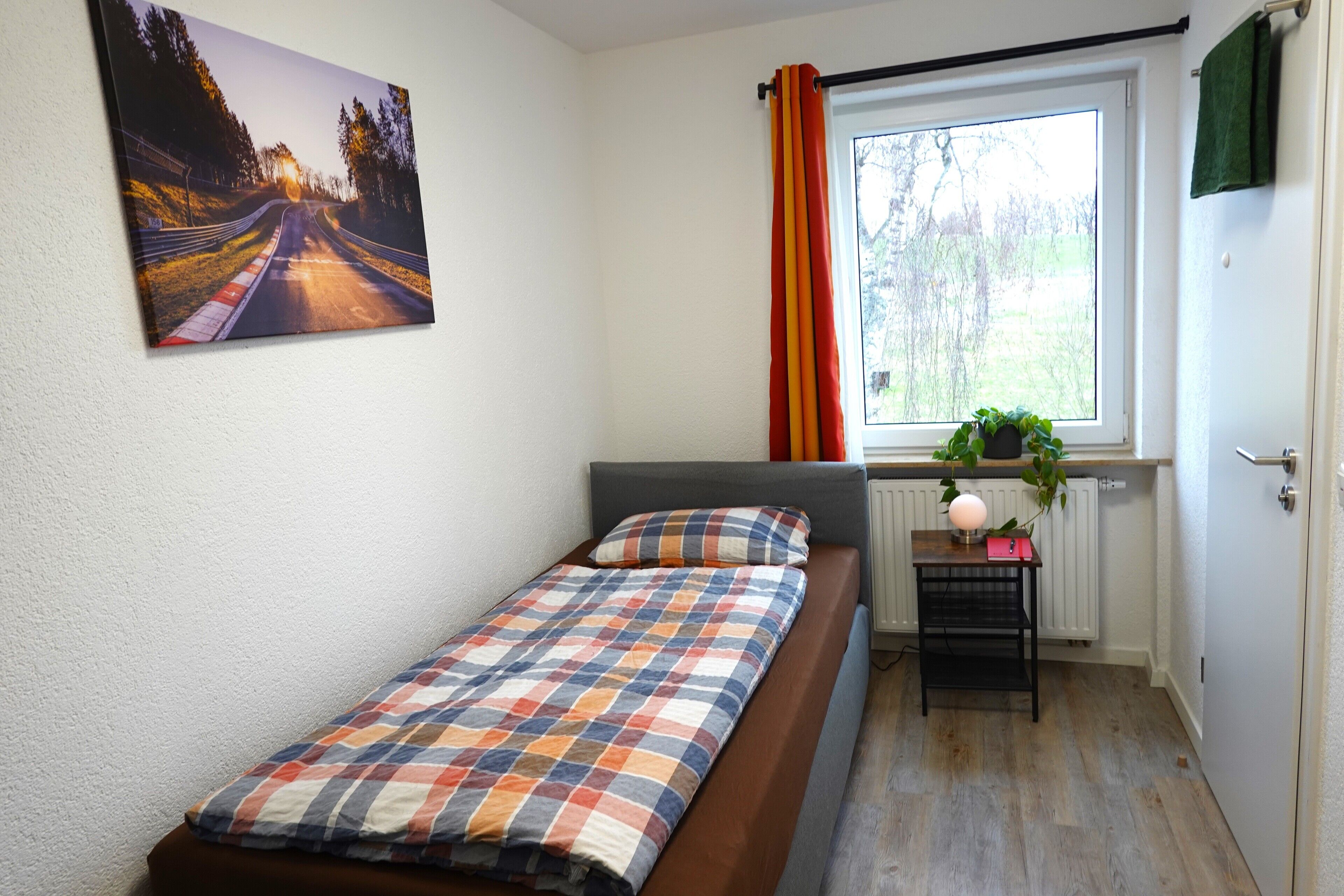 Quarto pequeno com cama de solteiro, janela, radiador, mesa de cabeceira com abajur e um quadro na parede.