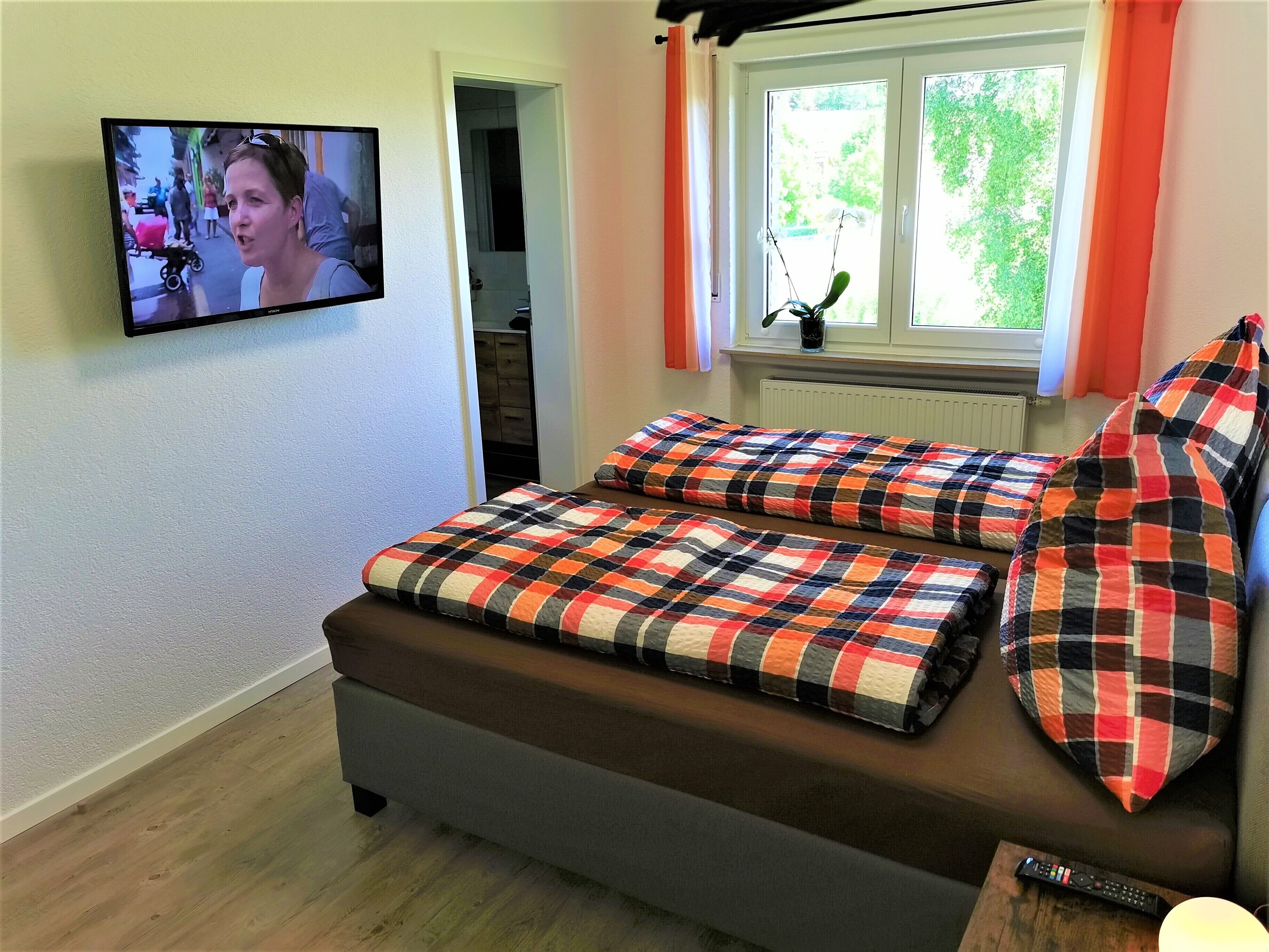 Um quarto com cama, televisão na parede, janela com cortinas e controle remoto sobre uma mesa de madeira.