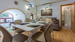 Uma sala de jantar com uma mesa branca posta para quatro pessoas, rodeada por cadeiras bege. Há um sofá e uma porta que dá para uma área de chuveiro.