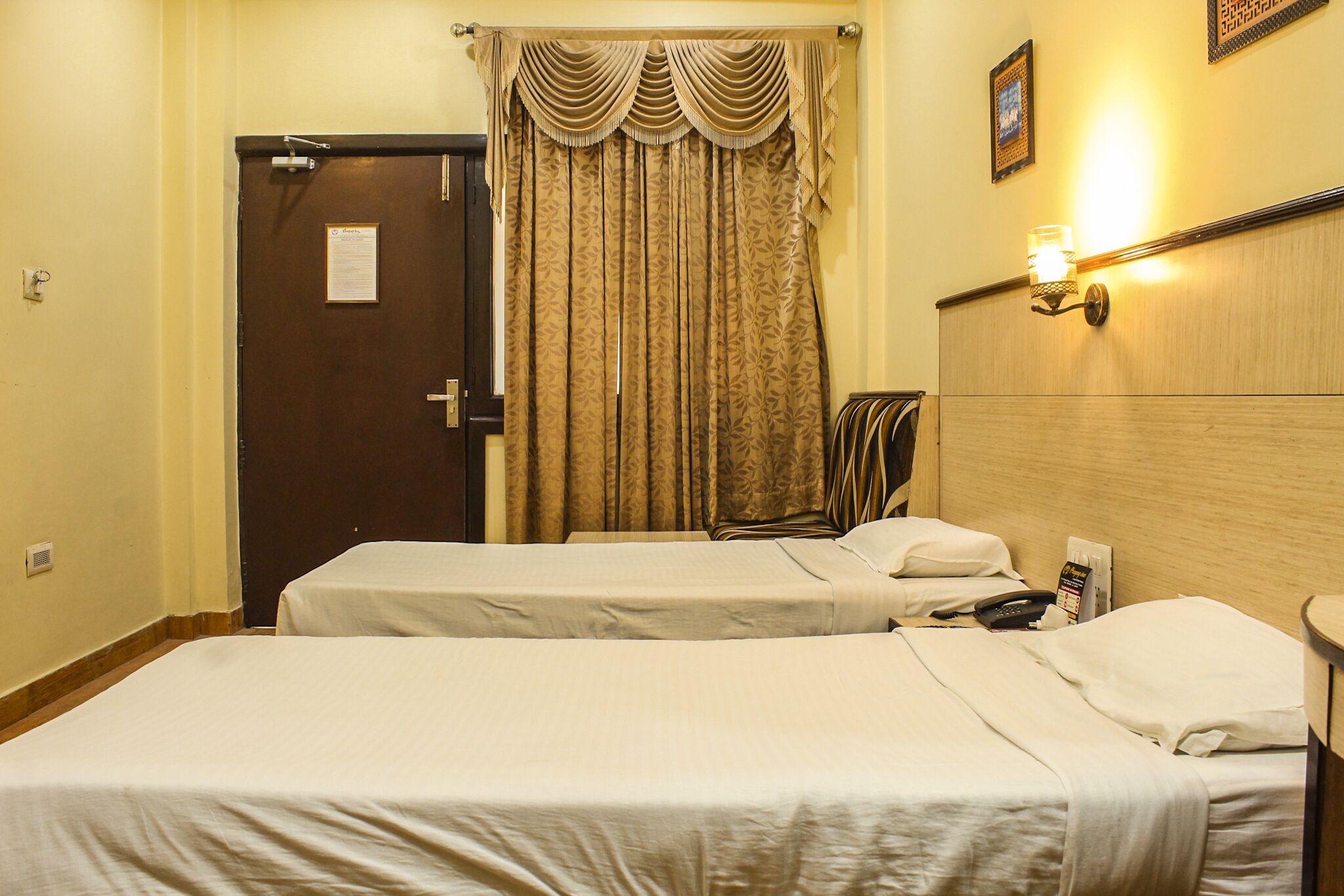 Quarto de hotel com duas camas, uma porta, uma cortina, uma luminária na parede e uma mesa de cabeceira com telefone.