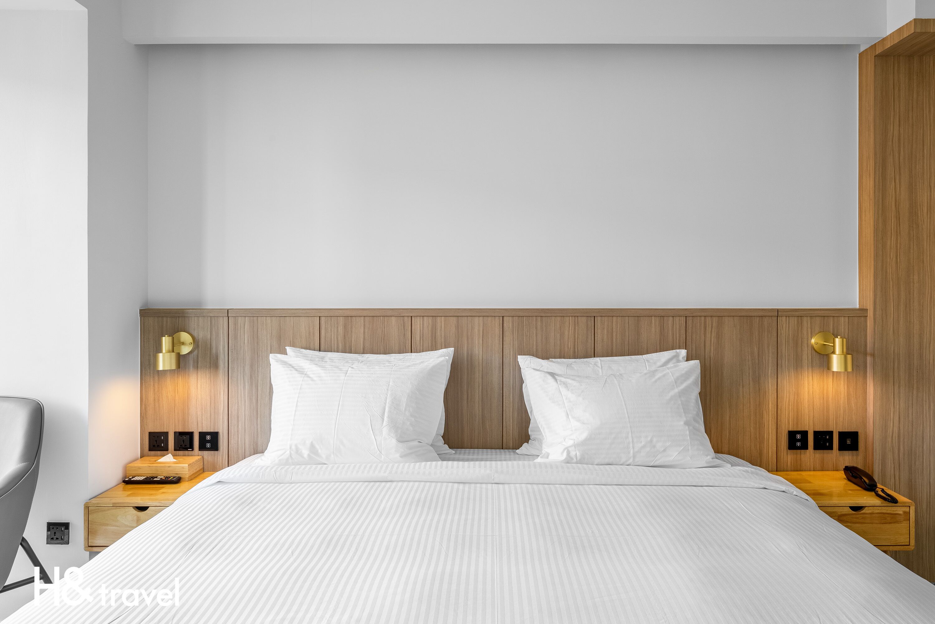 Quarto de hotel moderno com cabeceira de madeira, duas mesas de cabeceira com luminárias, cama com roupa de cama branca e uma cadeira cinza.