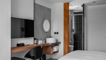 Quarto de hotel moderno com uma cama grande, uma escrivaninha de madeira com uma cadeira, televisão de tela plana, pia embutida e área de chuveiro.