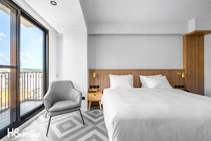 Quarto de hotel moderno com uma cama grande, uma poltrona cinza, uma mesa de cabeceira de madeira e vista para o exterior.