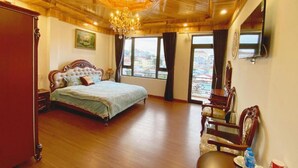 Um quarto espaçoso com uma cama grande, piso de madeira, um lustre, vista da cidade através de janelas amplas e uma televisão na parede.
