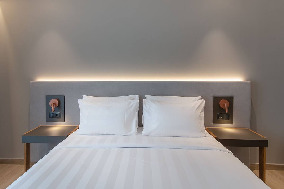 Quarto de hotel moderno com uma cama grande, duas mesas de cabeceira e iluminação embutida na parede.