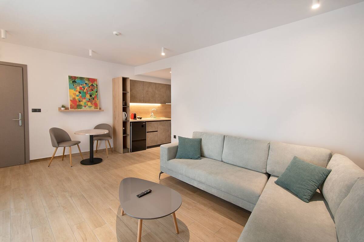 Uma sala de estar moderna com um sofá, uma mesa de centro e duas cadeiras. É visível uma área de cozinha ao fundo, com uma pia e armários.