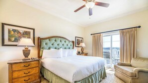 Quarto de hotel com cama grande, cômoda de madeira, duas luminárias de cabeceira, ventilador de teto e varanda com cortinas.