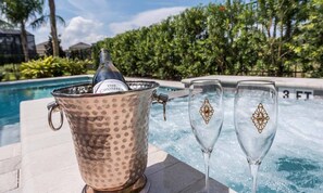 Ambiente à beira da piscina, com um balde de gelo de cobre contendo uma garrafa com a etiqueta "The Crusher", duas taças elaboradas e uma piscina com paisagem tropical.