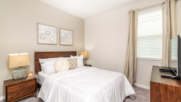 Quarto com cama grande, duas mesas de cabeceira com luminárias, criado-mudo de madeira, cômoda e janela com cortinas.