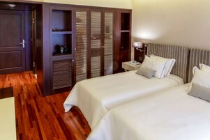 Quarto de hotel com duas camas, um guarda-roupa de madeira, uma mesa de cabeceira com abajur e cabeceira listrada.
