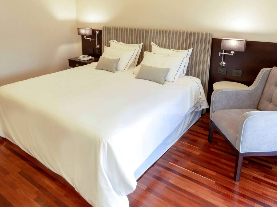 Quarto de hotel com uma cama grande, duas mesas de cabeceira com luminárias, uma cadeira e piso de madeira.