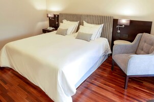 Quarto de hotel com uma cama grande, duas mesas de cabeceira com luminárias, uma cadeira e piso de madeira.