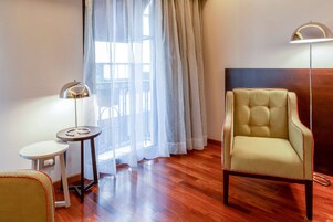 Um quarto com piso de madeira, uma poltrona bege, uma pequena mesa branca, um abajur e cortinas leves.