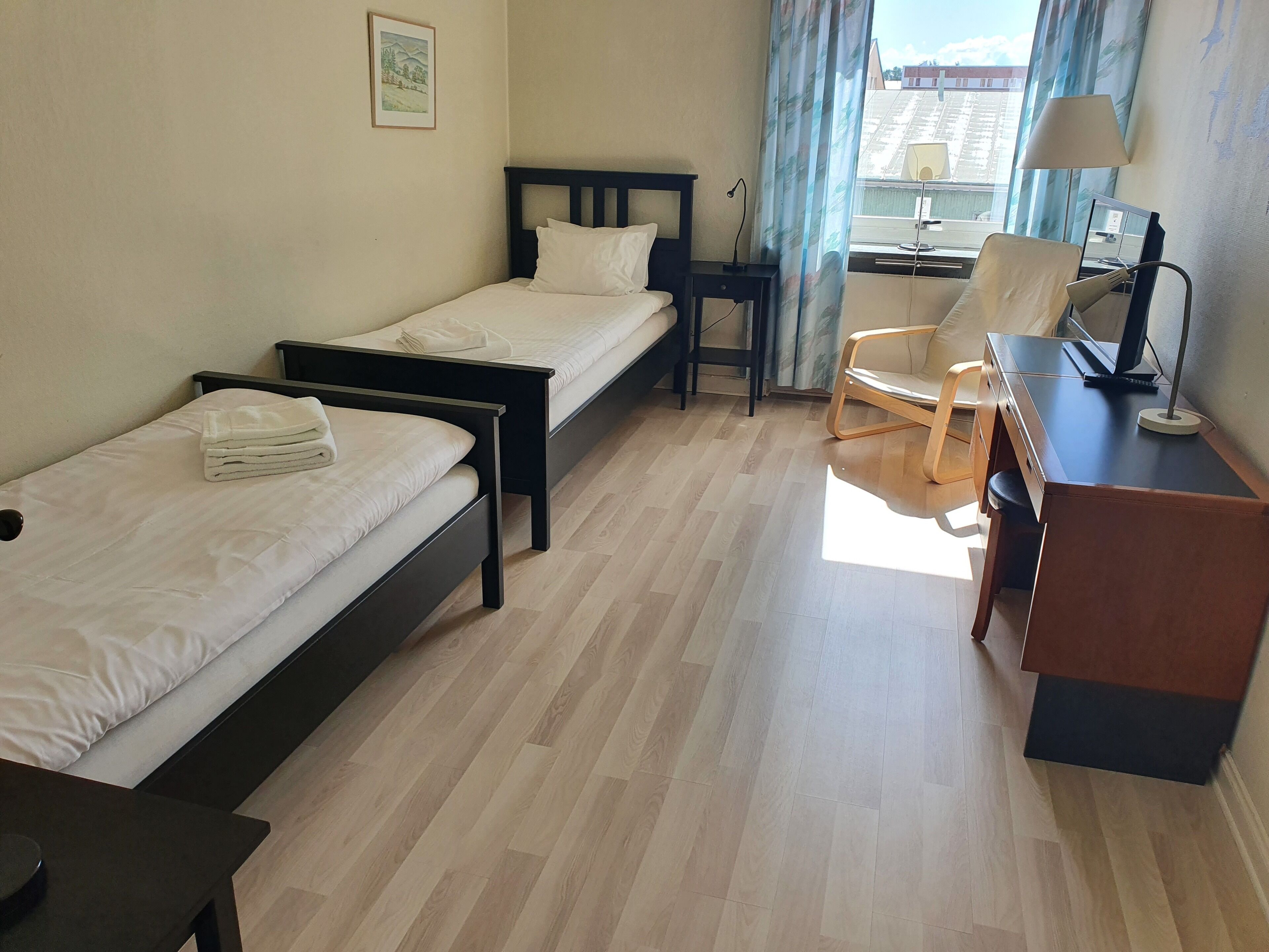 Quarto de hotel com duas camas de solteiro, uma mesa com cadeira, um abajur e uma janela com cortinas.