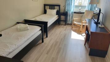 Quarto de hotel com duas camas de solteiro, uma mesa com cadeira, um abajur e uma janela com cortinas.