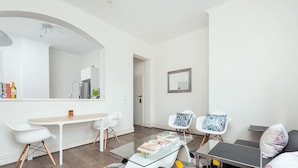 Quarto moderno e bem iluminado, com mesa de vidro, duas cadeiras brancas, um sofá e uma mesinha de centro.