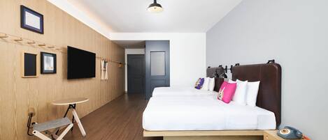 Quarto de hotel moderno com piso de madeira, cama com roupa de cama branca, mesa de cabeceira com telefone e pequena escrivaninha com cadeira.