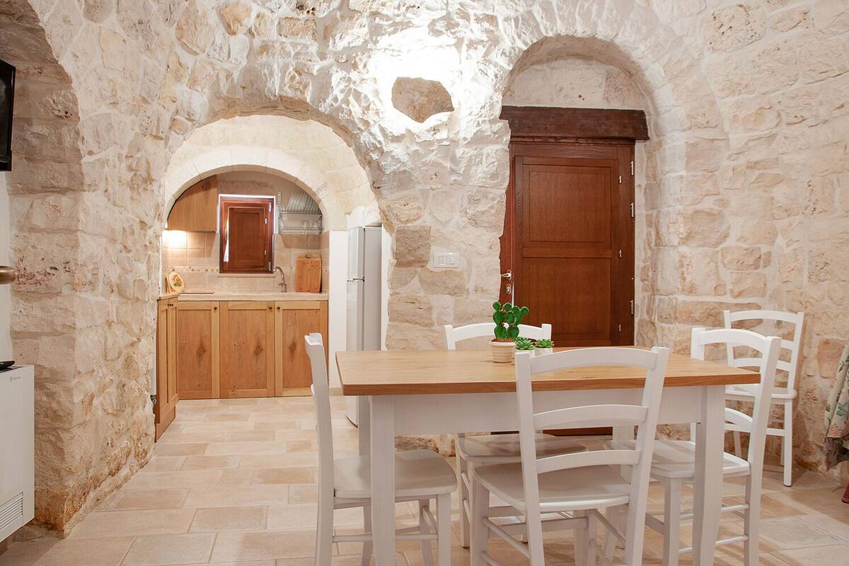 Um quarto com paredes de pedra, uma mesa e cadeiras de madeira, um espaço de cozinha com armários de madeira e uma geladeira, e uma grande porta de madeira.