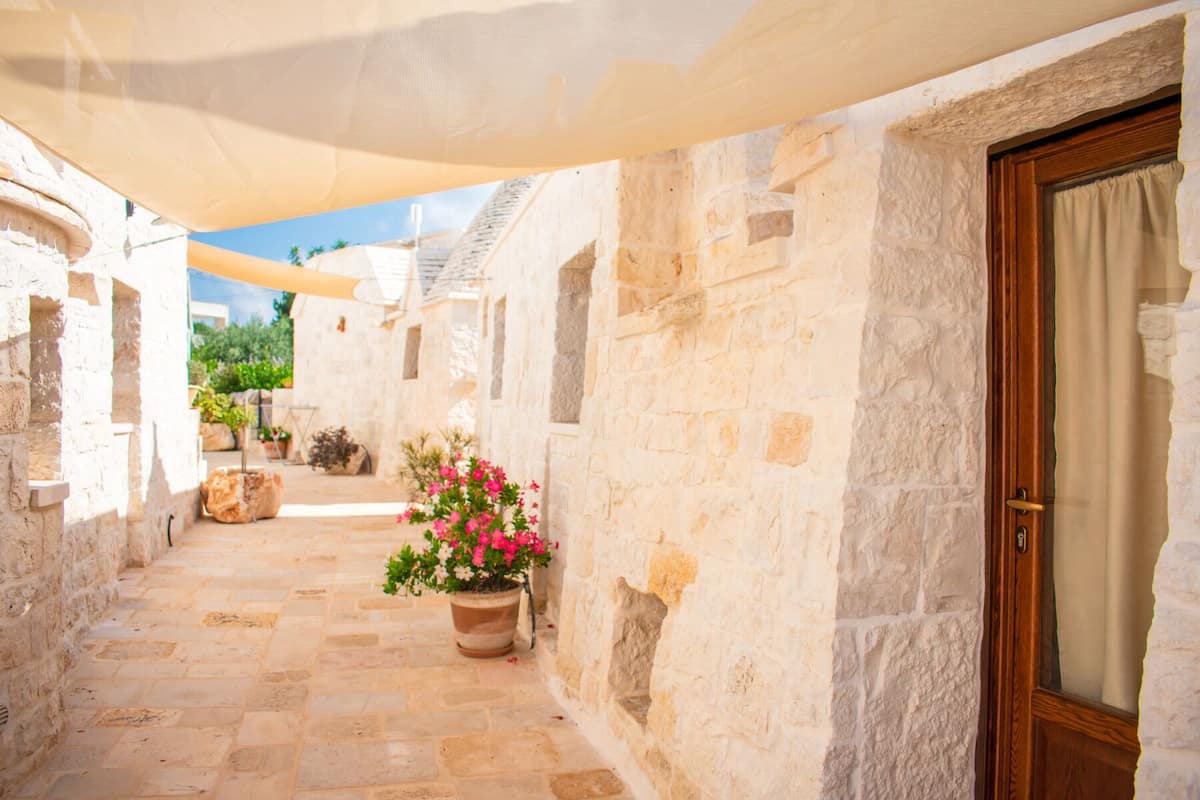 Um caminho de pedra estreito que leva a um edifício com porta de madeira, ladeado por plantas em vasos e uma área sombreada.