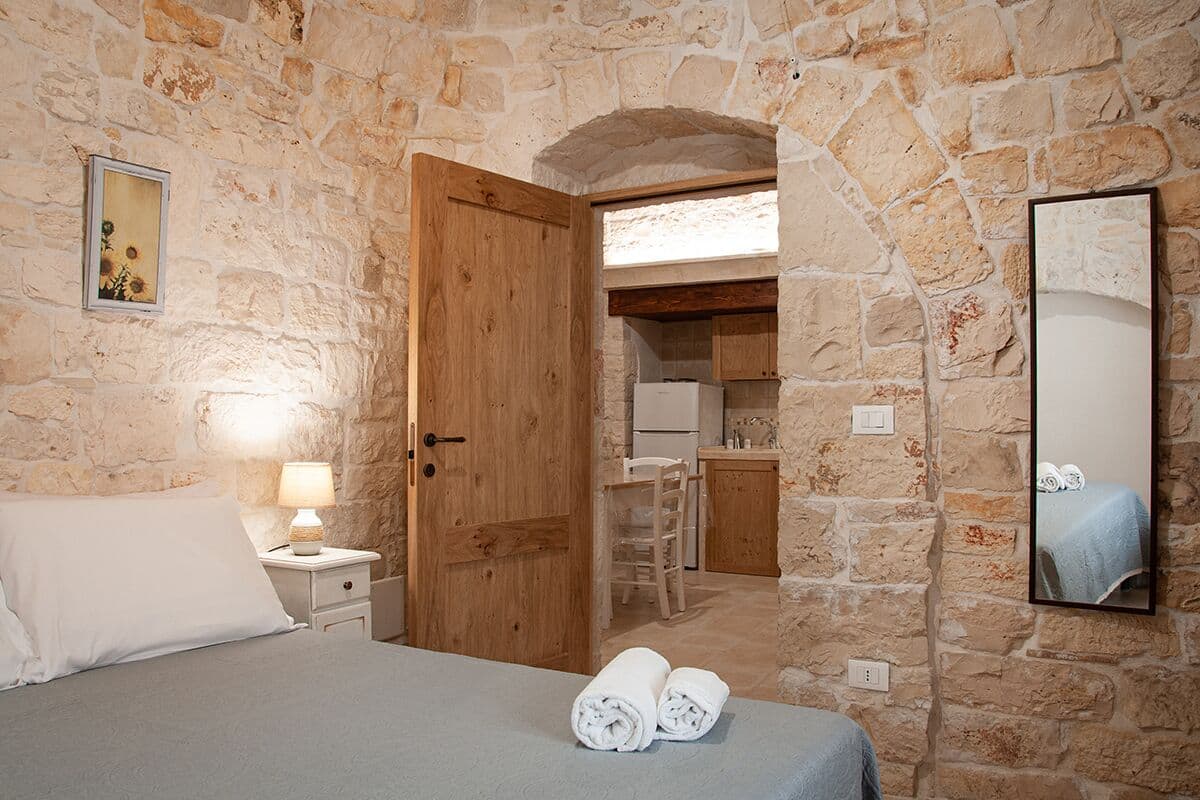 Quarto com parede de pedra, porta de madeira, cama com roupa de cama branca, mesa de cabeceira com abajur e espelho na parede.