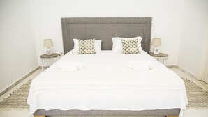 Cama bem arrumada com cabeceira cinza, duas mesas de cabeceira com luminárias e um tapete tecido no chão.