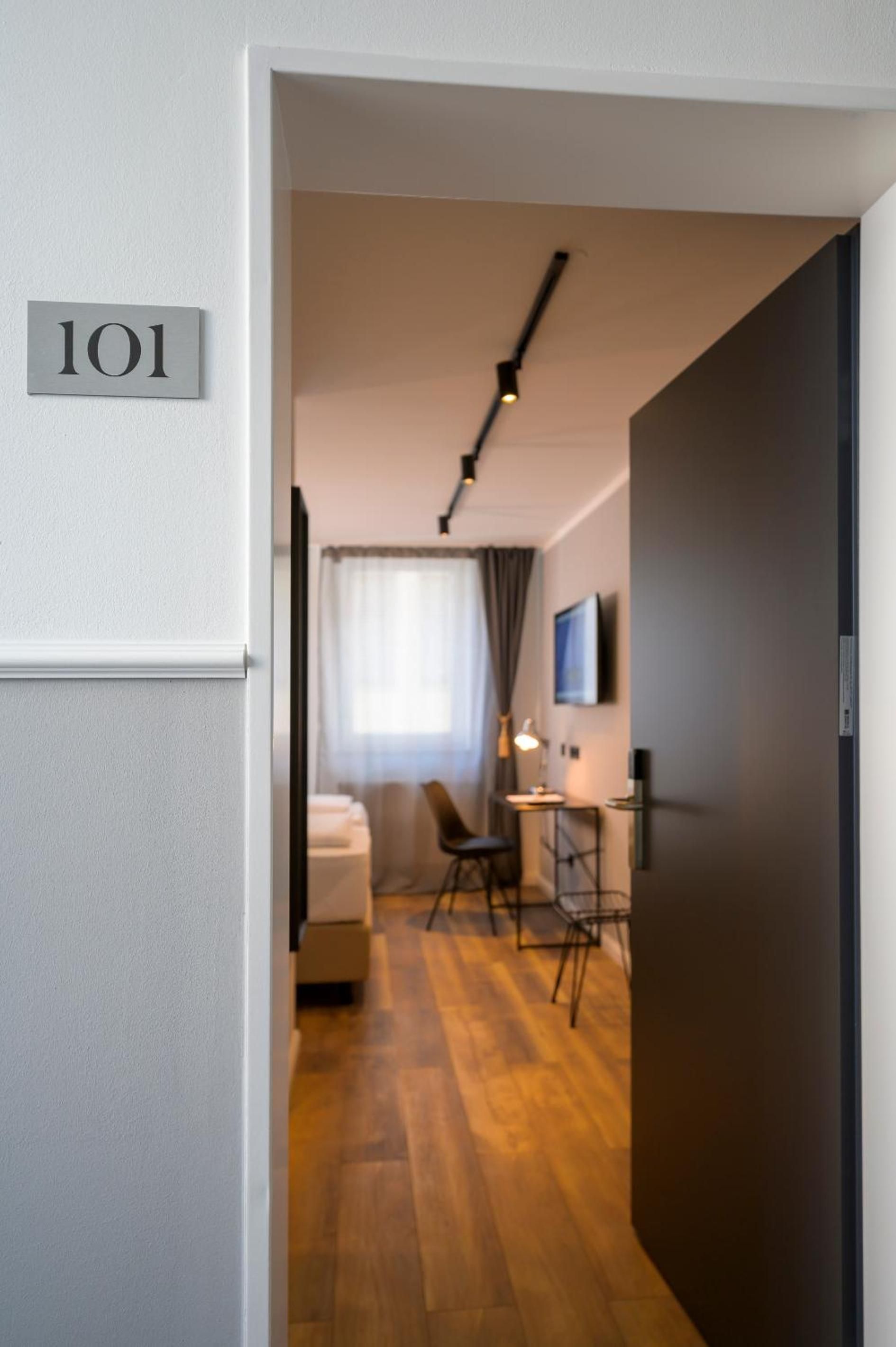 Corredor que dá para um quarto com placa indicando o número '101', uma cama, uma mesa com cadeira e uma TV fixada na parede.