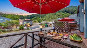 Uma área de refeições com uma mesa posta para refeição, um guarda-sol vermelho e vista para uma rua residencial.