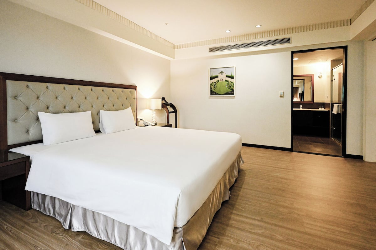 Quarto de hotel com uma cama grande, mesa de cabeceira, abajur, quadro na parede e banheiro visível através de uma porta aberta.
