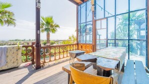 Um deck de madeira com mesa e cadeiras, uma área coberta com janelas amplas e vista para palmeiras e vegetação.