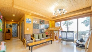 Quarto aconchegante com teto de madeira, um sofá com almofadas, um banco, um cercado infantil e vista para palmeiras lá fora.