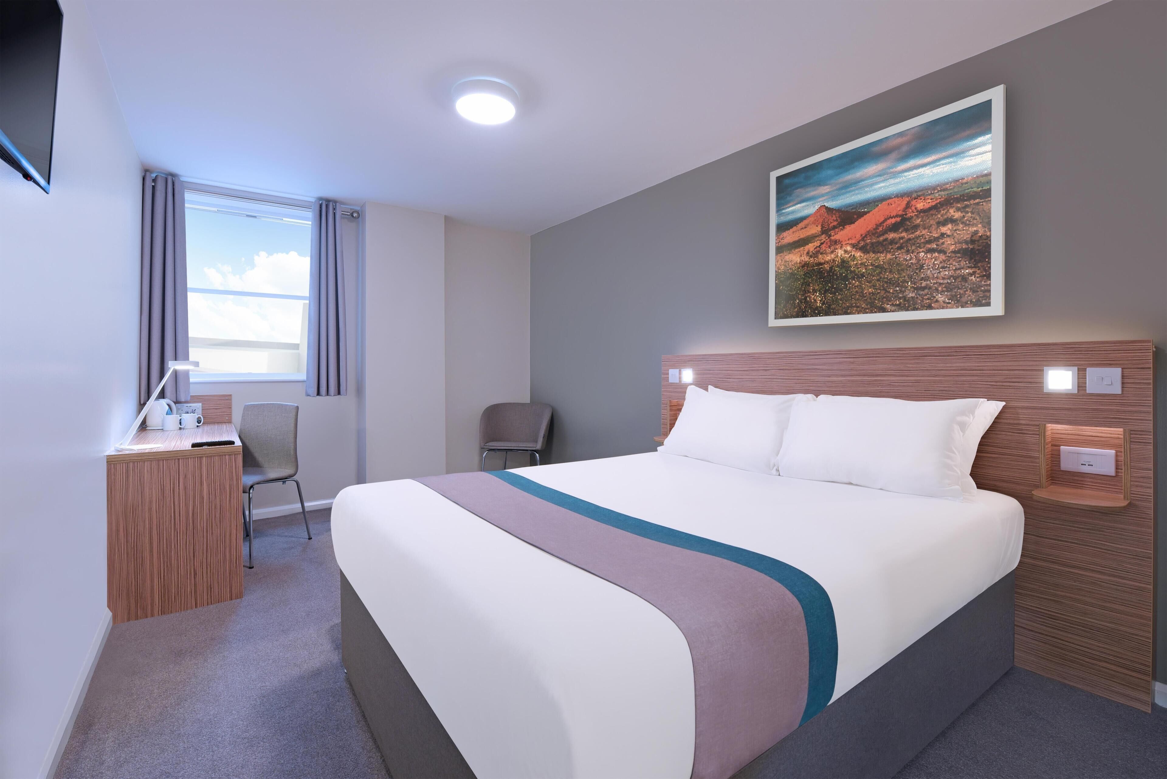 Quarto de hotel com cama, escrivaninha, cadeira e um quadro na parede.