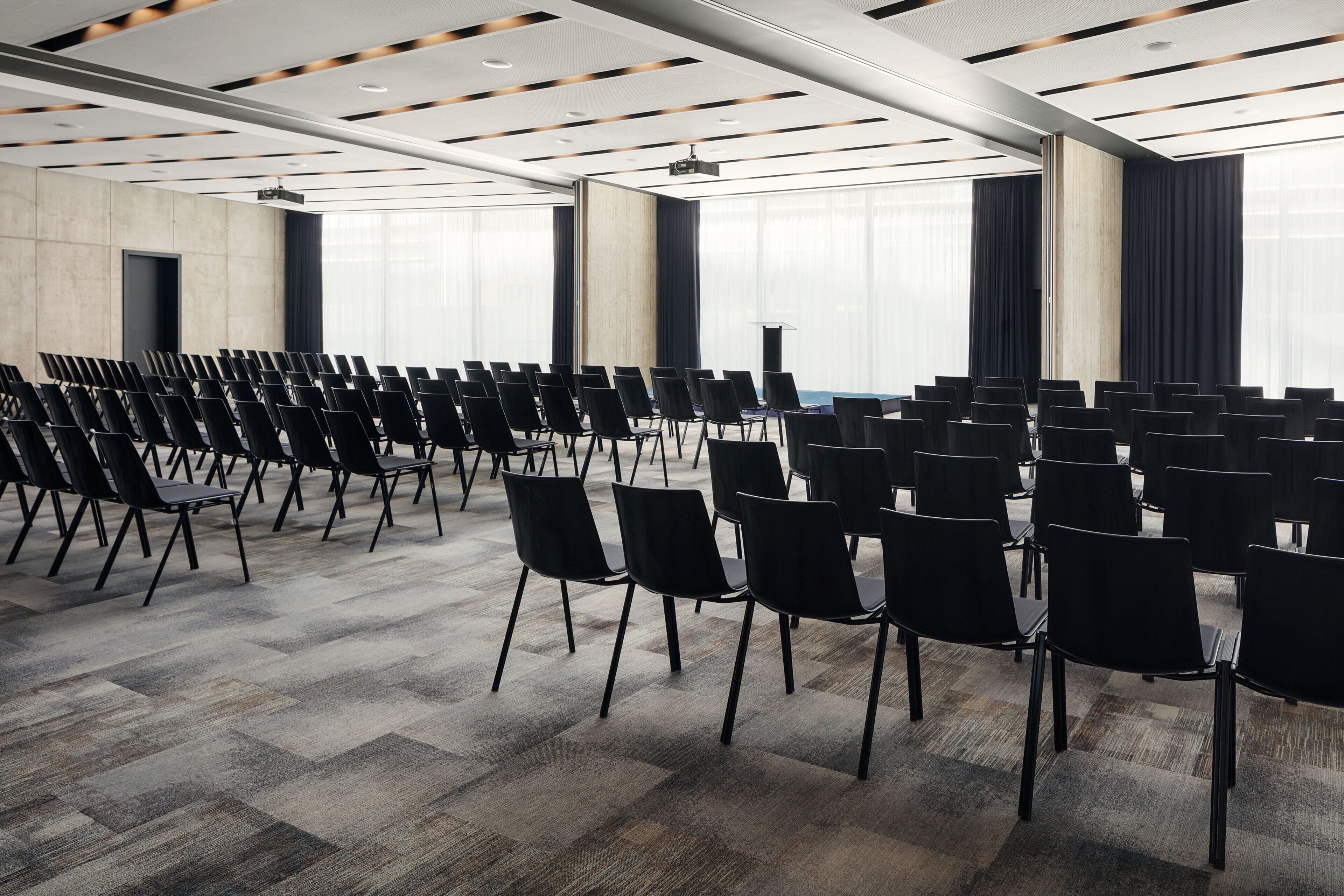 Uma sala de conferências com fileiras de cadeiras pretas, um projetor e uma janela grande com cortinas.