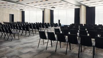 Uma sala de conferências com fileiras de cadeiras pretas, um projetor e uma janela grande com cortinas.