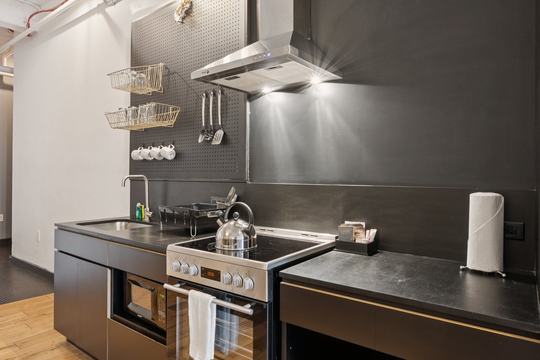 Uma cozinha moderna com eletrodomésticos de aço inoxidável, incluindo fogão, forno e coifa. Há utensílios pendurados e um porta-toalhas na parede.