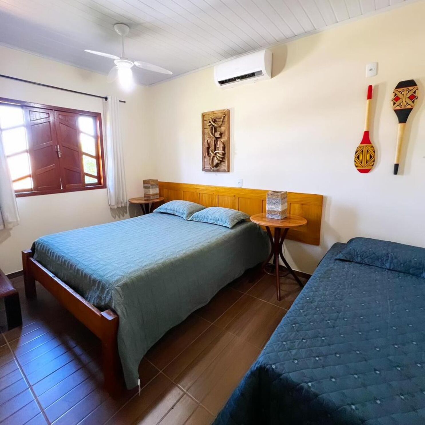 Quarto com duas camas, cabeceira de madeira, uma mesinha, ar condicionado fixado na parede e ventilador de teto.