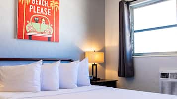 Quarto de hotel com cama, almofadas brancas, mesa de cabeceira com abajur, janela com cortinas e um letreiro na parede com a inscrição "BEACH, PLEASE".